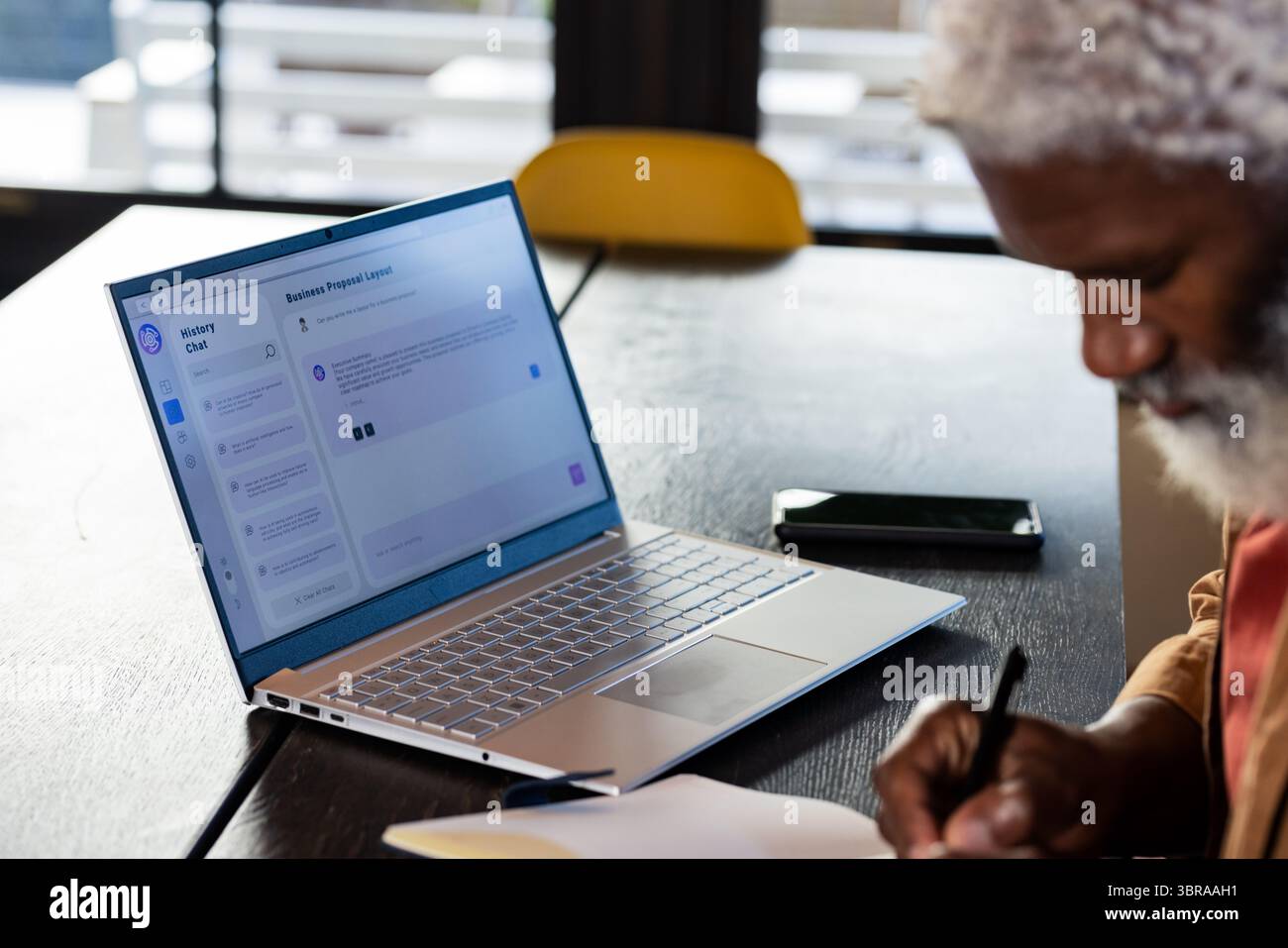 Senior African American man jotting notes at office desk beside laptop ...