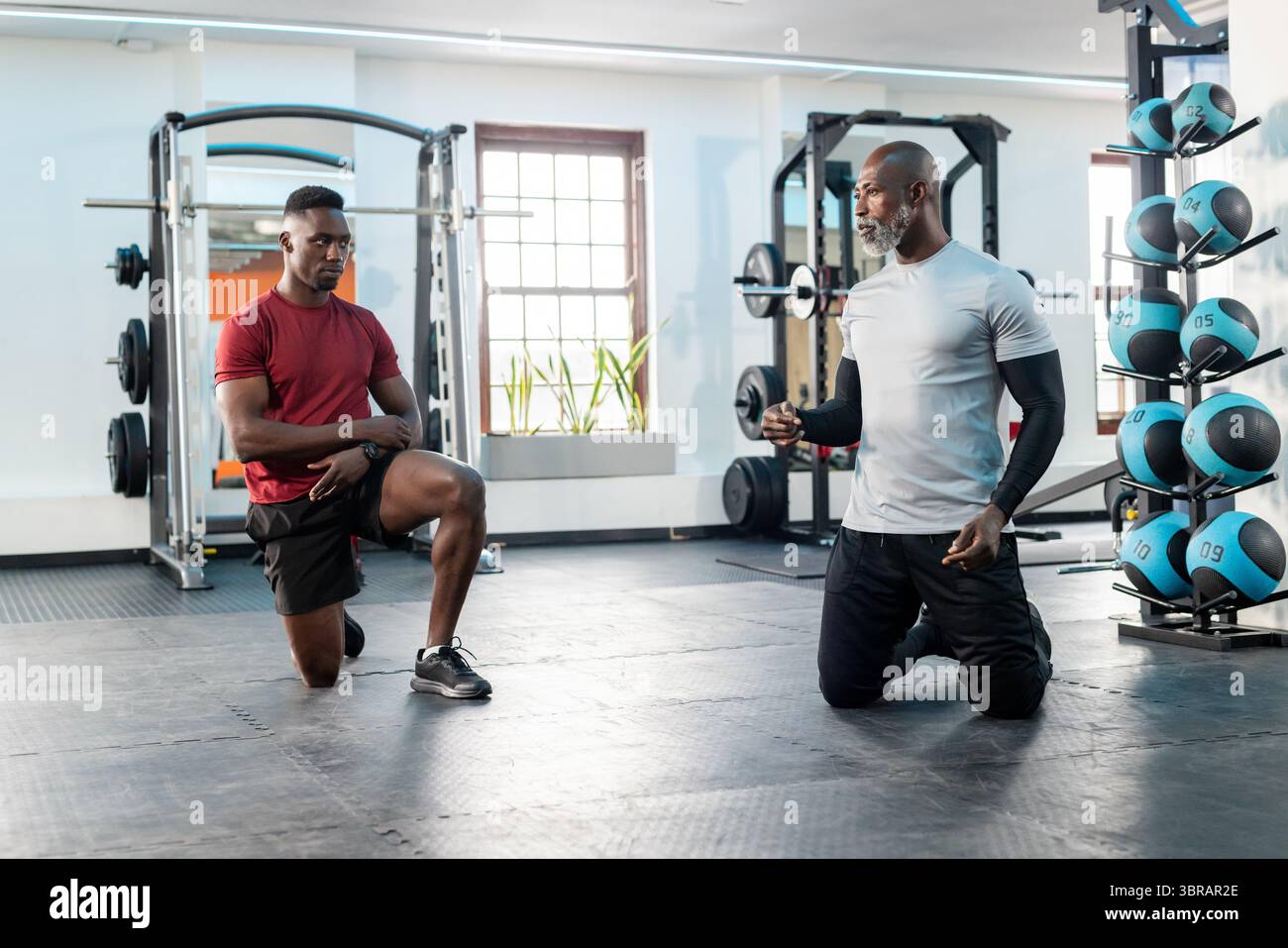 African American male trainer and trainee kneeling at gym, improving ...