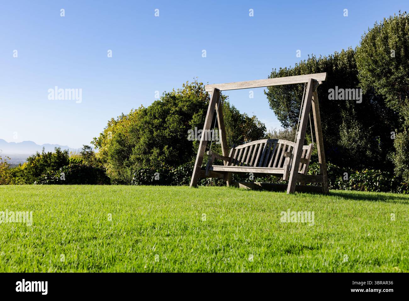 wooden swing bench is resting on green lawn under clear blue sky, shrub ...