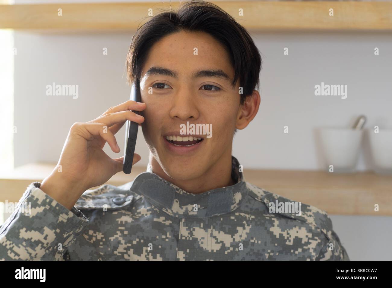 Asian male soldier speaking on smartphone in kitchen with wooden ...