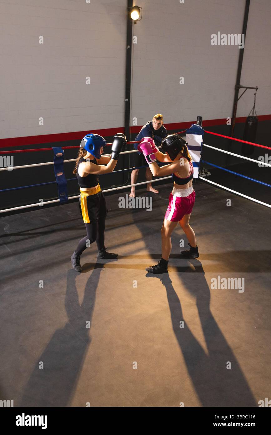 Two female boxers sparring inside boxing ring at gym with headgear, boxing gloves, punching bag ...