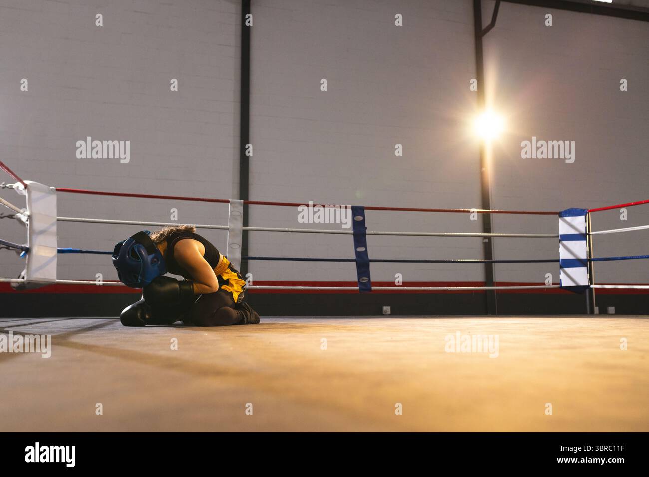 female boxer kneeling on ring mat under spotlight wearing blue headgear ...
