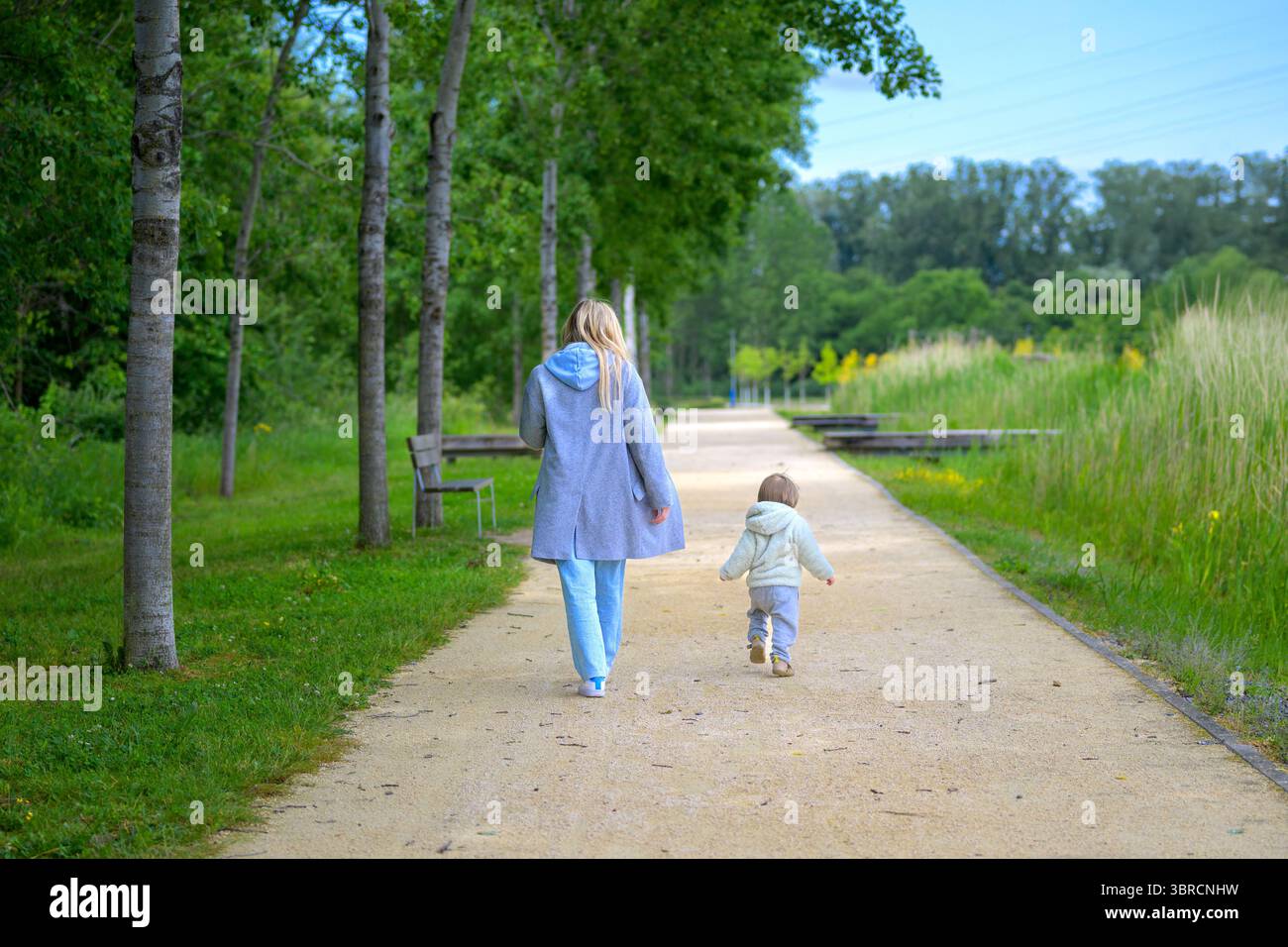 A blonde woman and a toddler walk side by side down a wide park path ...