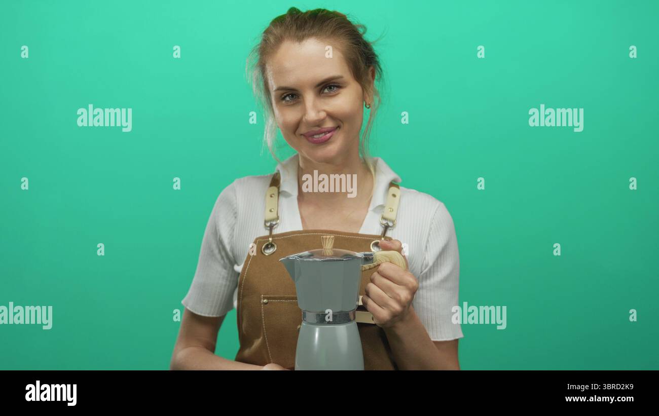 Woman smiling in apron holding coffee maker against green background ...