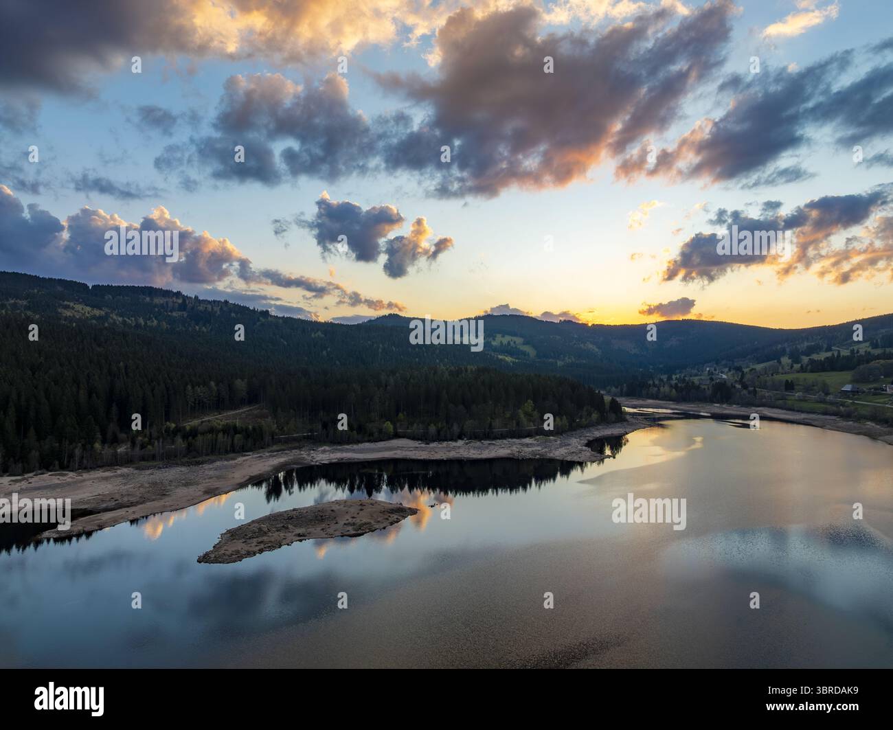 Aerial view of a serene lake reflecting the pastel sunset sky, nestled between dark forests and ...