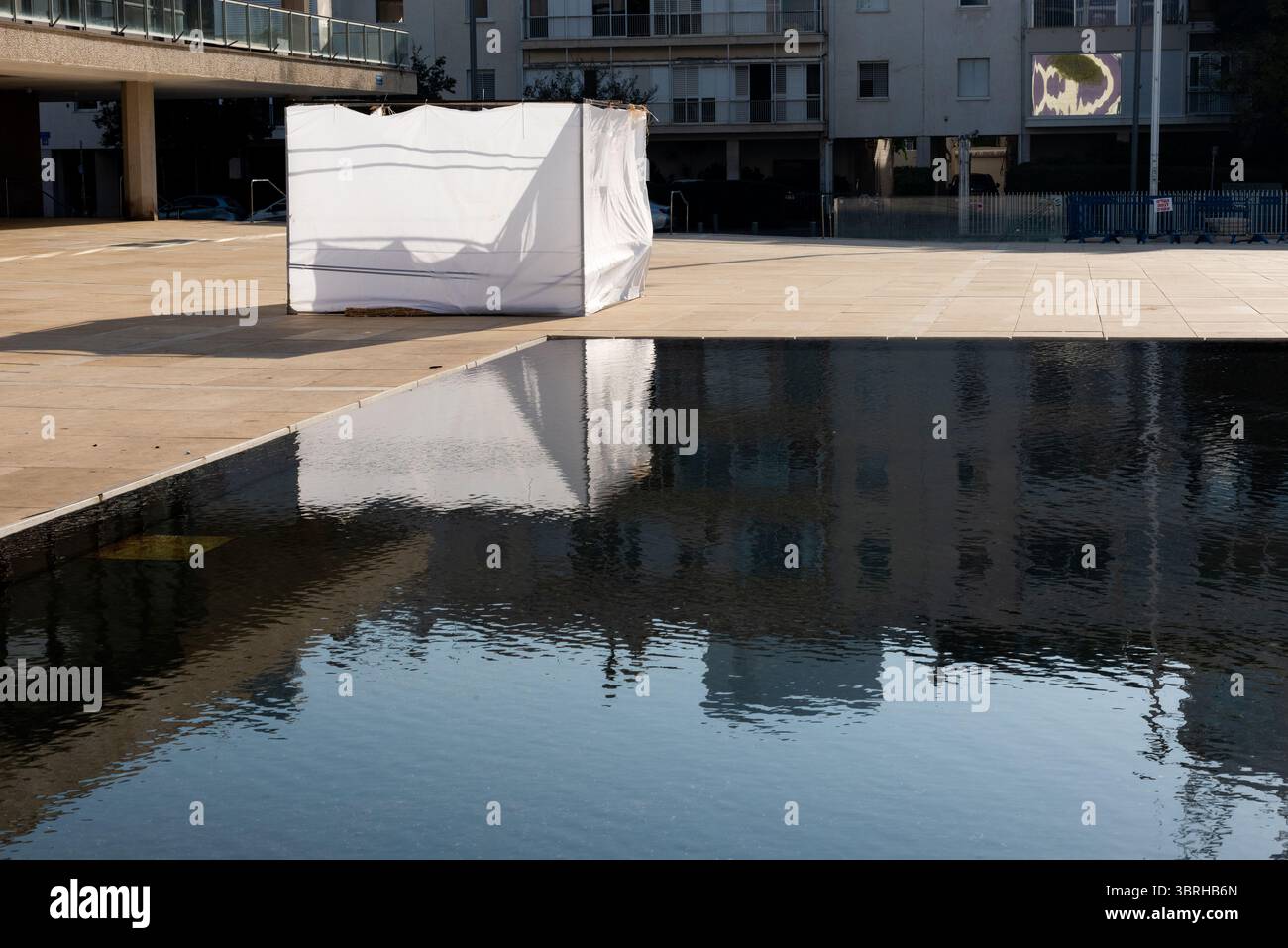 A single, white fabric sukkah booth reflected in a small pool of water ...