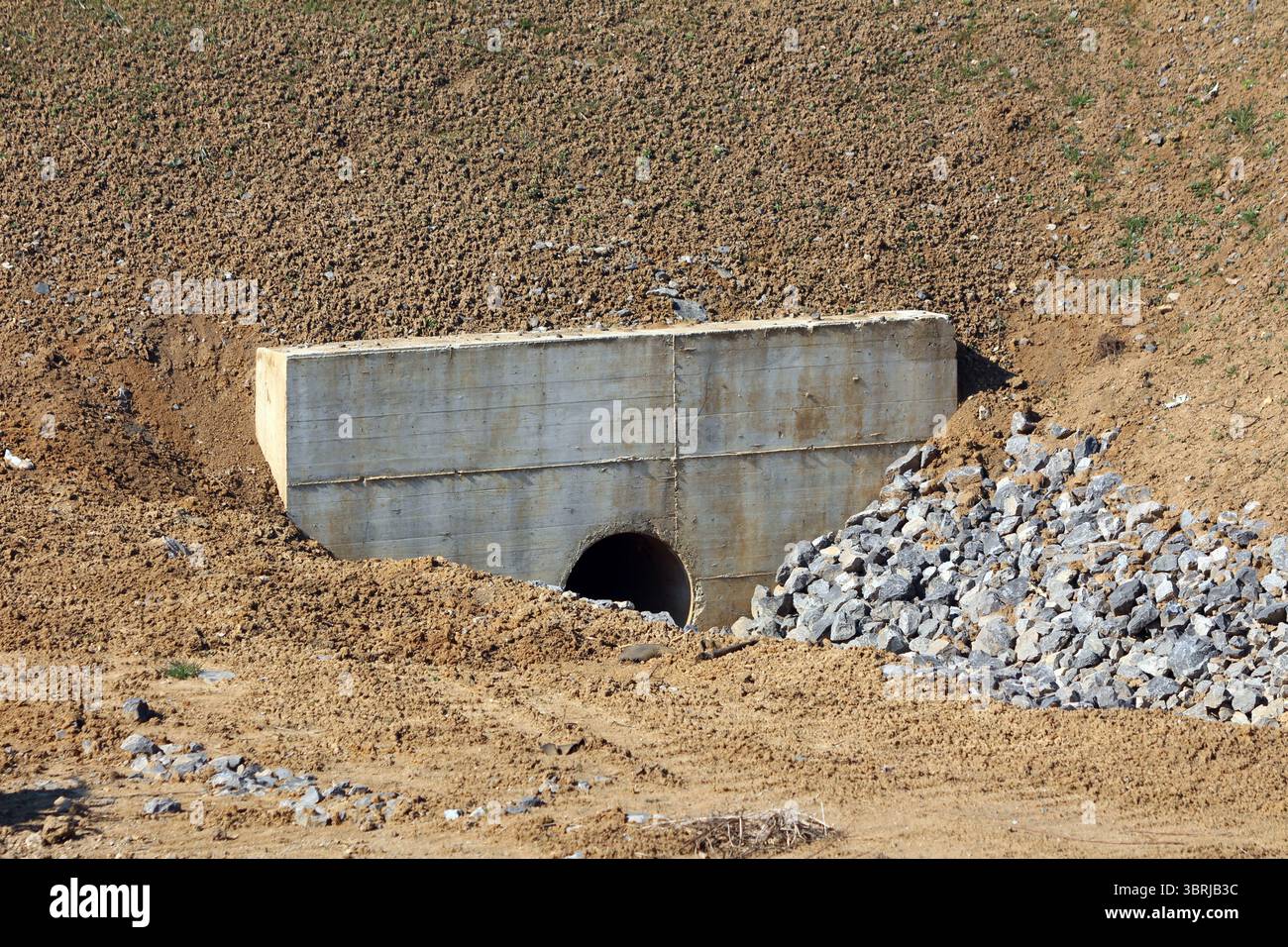 A reinforced concrete culvert pipe emerges from a sloped, eroded earth ...