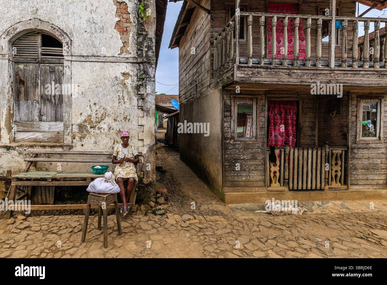 a local villager sits on a wooden bench in front of a rundown colonial ...