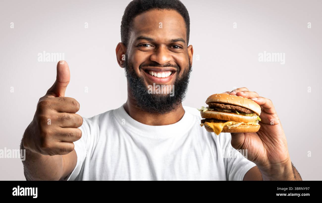 Black Man Holding Burger And Gesturing Thumbs-Up Over Yellow Background ...
