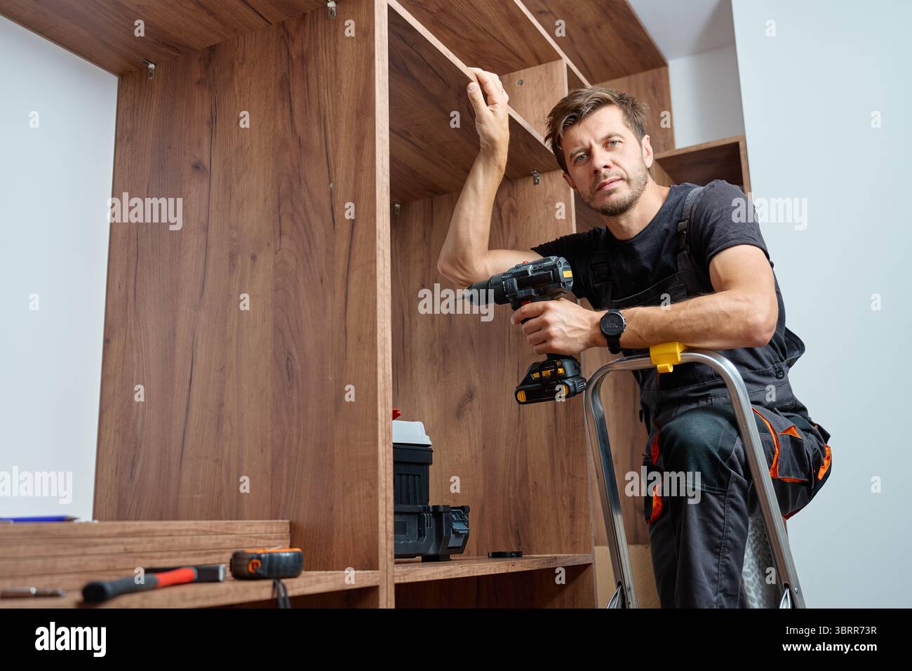 Man wearing work overalls assembling wooden wardrobe with drill and tools. Carpenter standing on ...