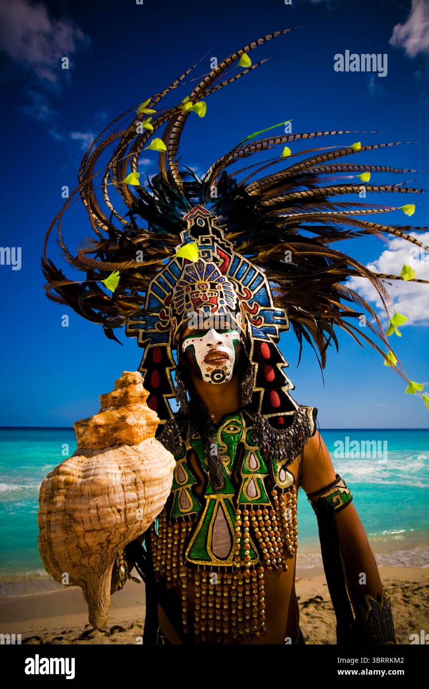 A man dressed in an elaborate headdress and painted face offers a conch ...