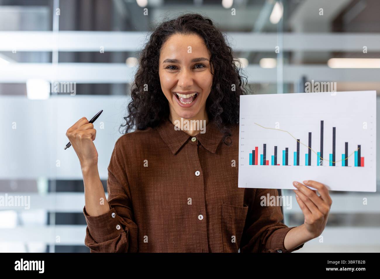A happy woman in a brown shirt holding a chart with a graph and a pen ...