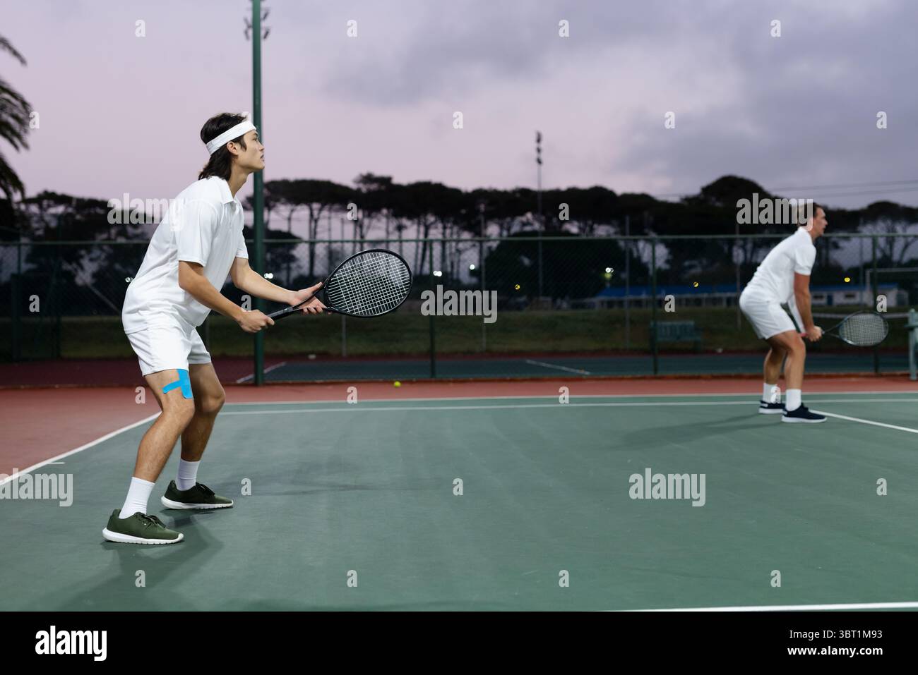 Diverse male doubles partners standing ready on tennis court at dusk ...