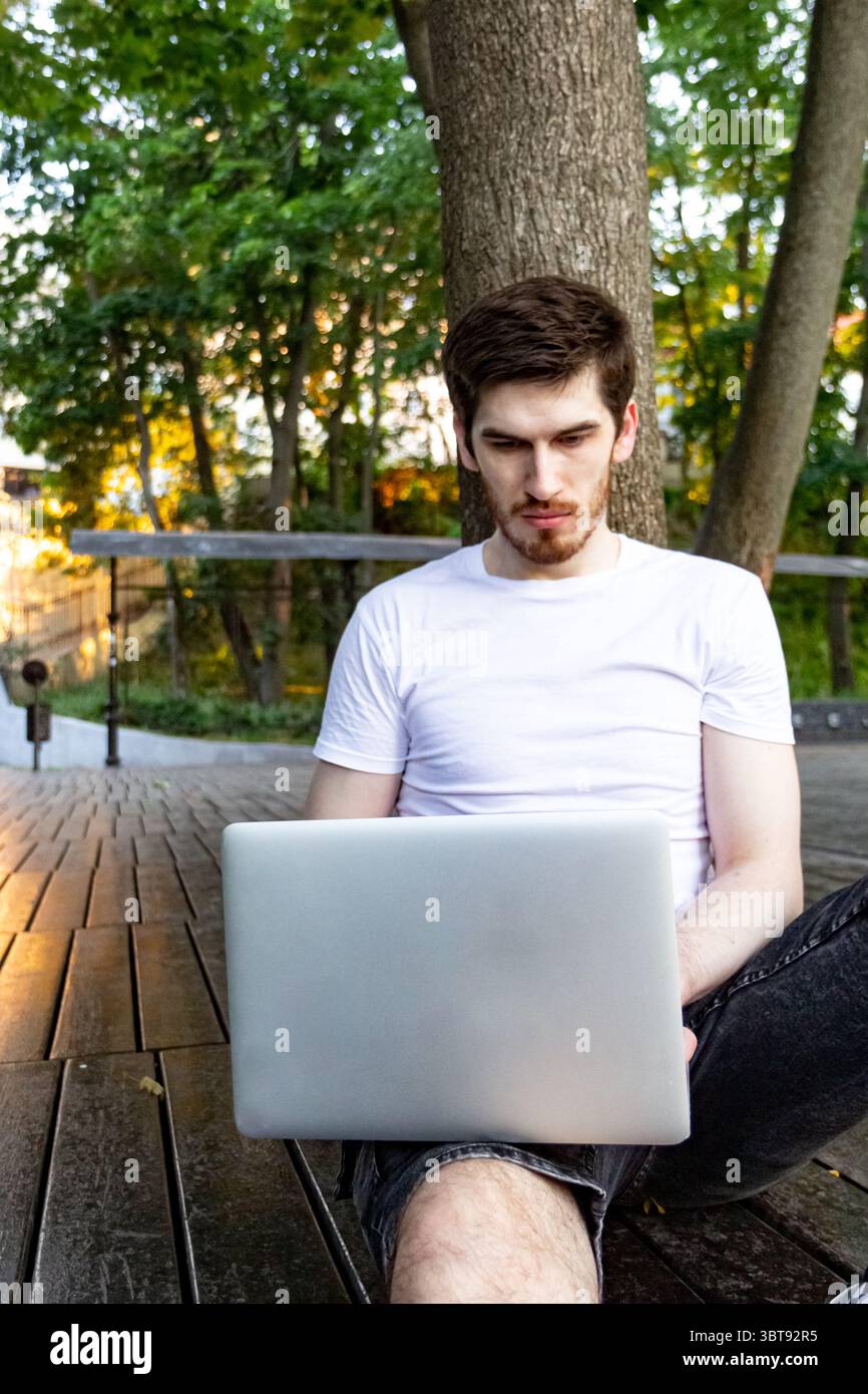 Casual man sits on wooden deck, back against tree, typing on laptop ...