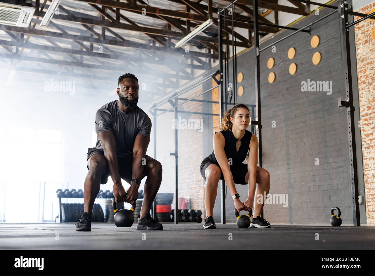 Diverse training partners wearing sportswear lifting kettlebells in squat stance in CrossFit gym ...