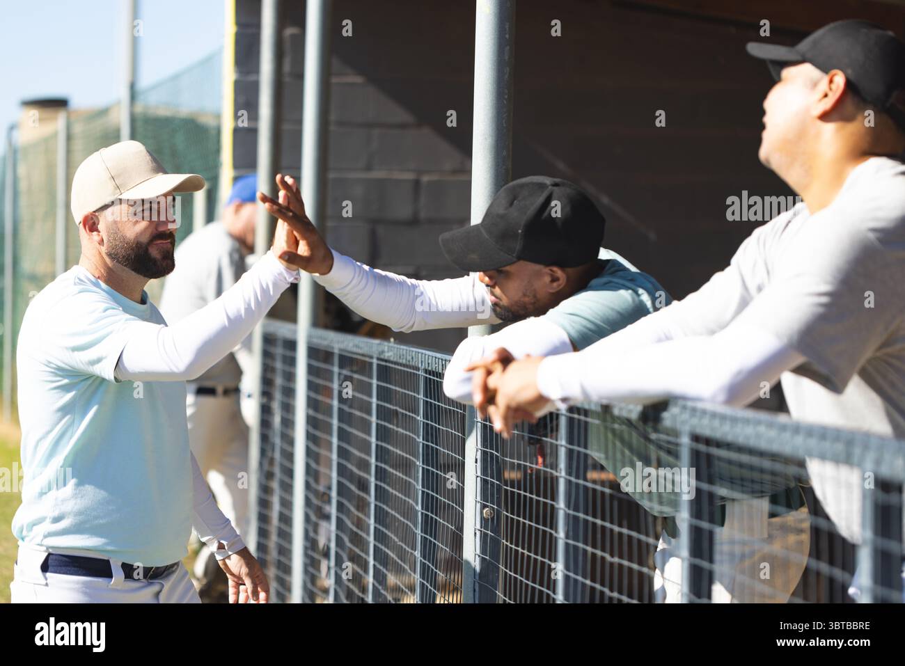 Diverse male baseball players exchanging high-five by chain-link fence ...