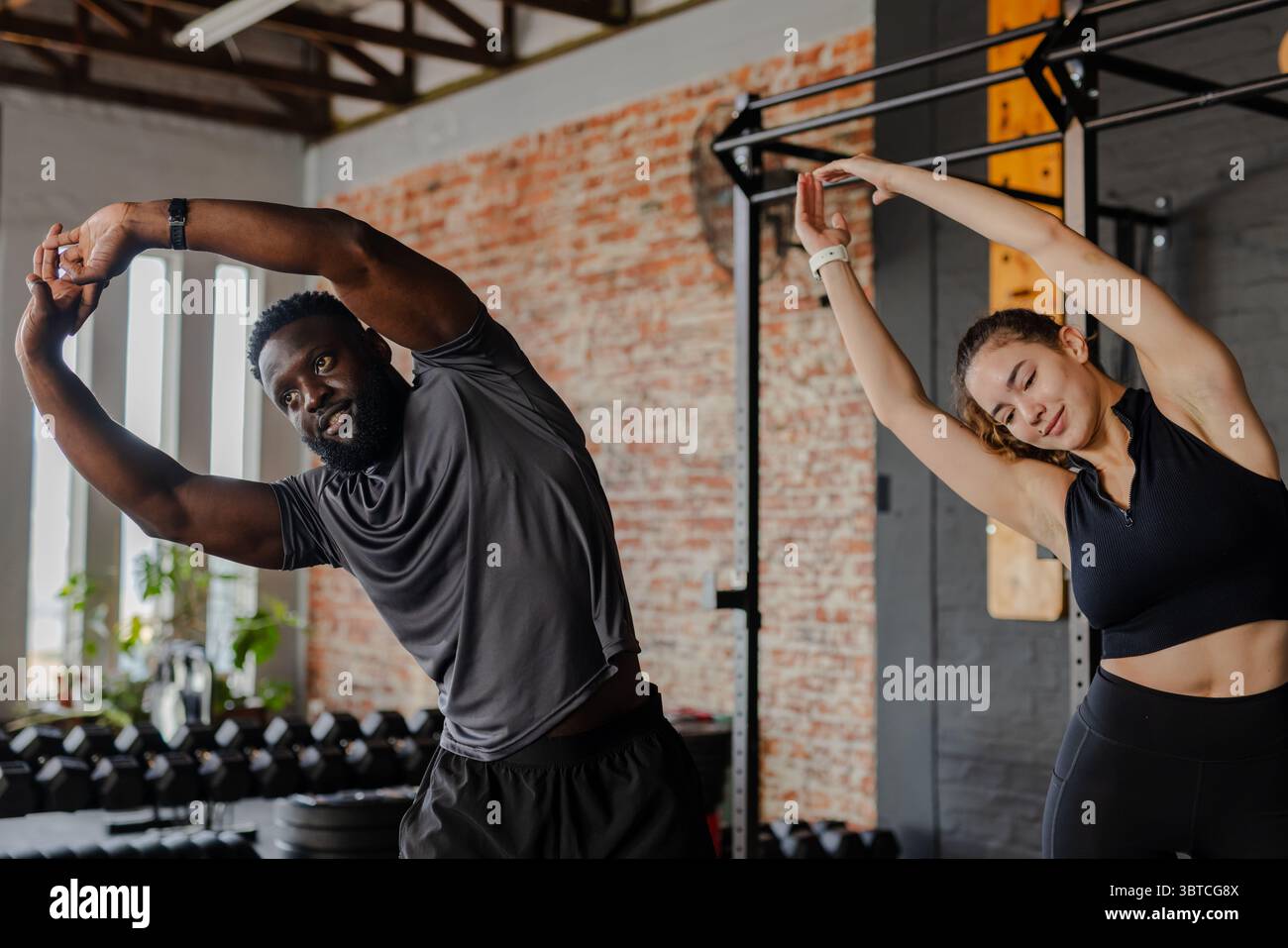 Diverse workout partners stretching sideways at gym near dumbbell rack ...