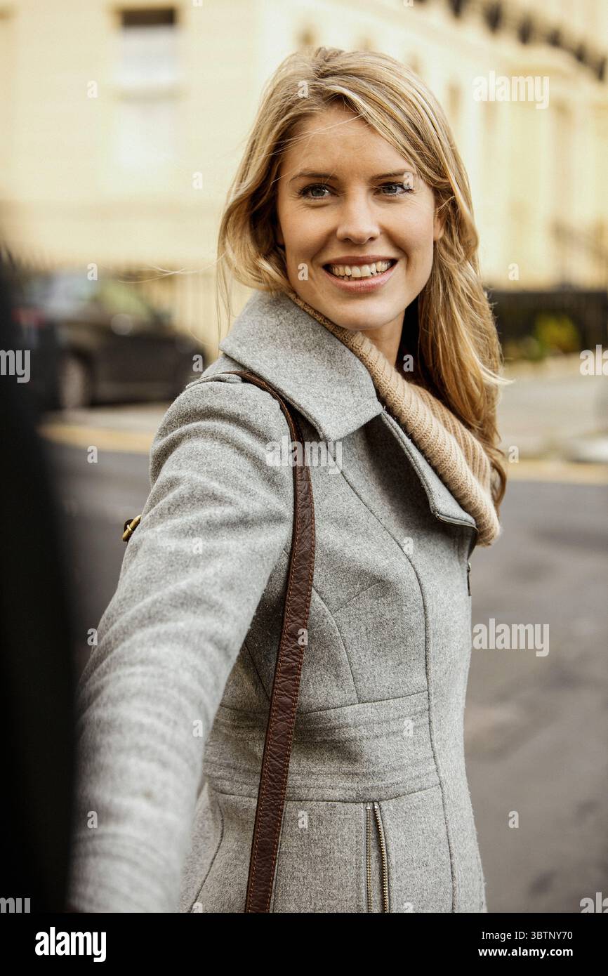 Beautiful blond woman in gray coat turning and smiling on sidewalk ...