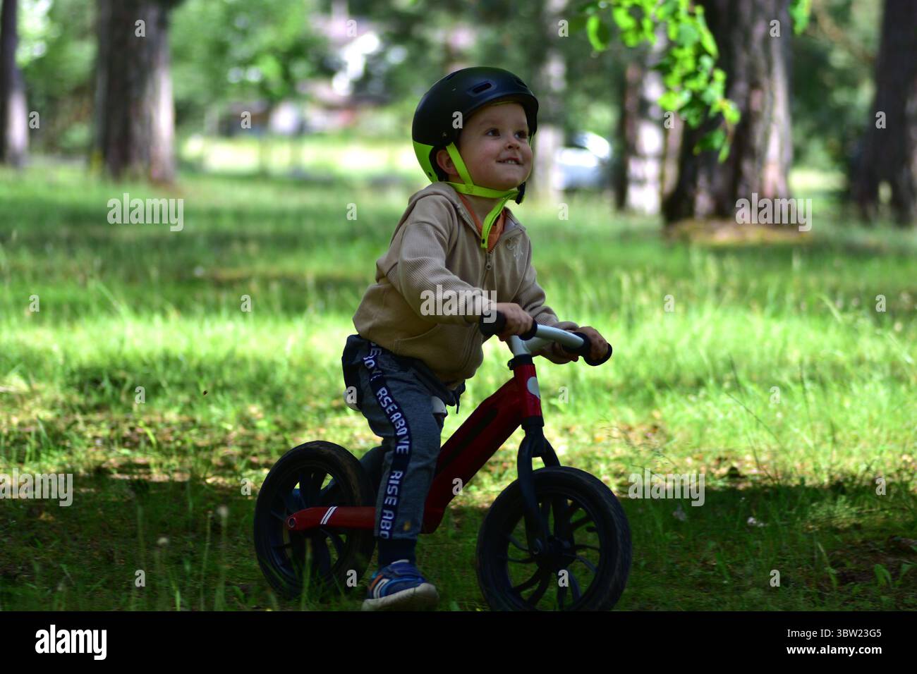 Happy boy riding red balance bike in green park, looking up with a big smile. Scene captures ...