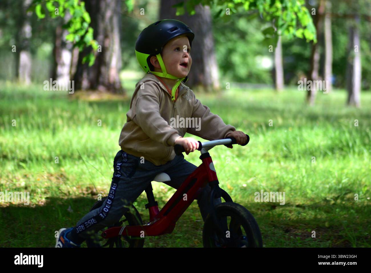 Happy boy riding red balance bike in green park, looking up with a big smile. Scene captures ...