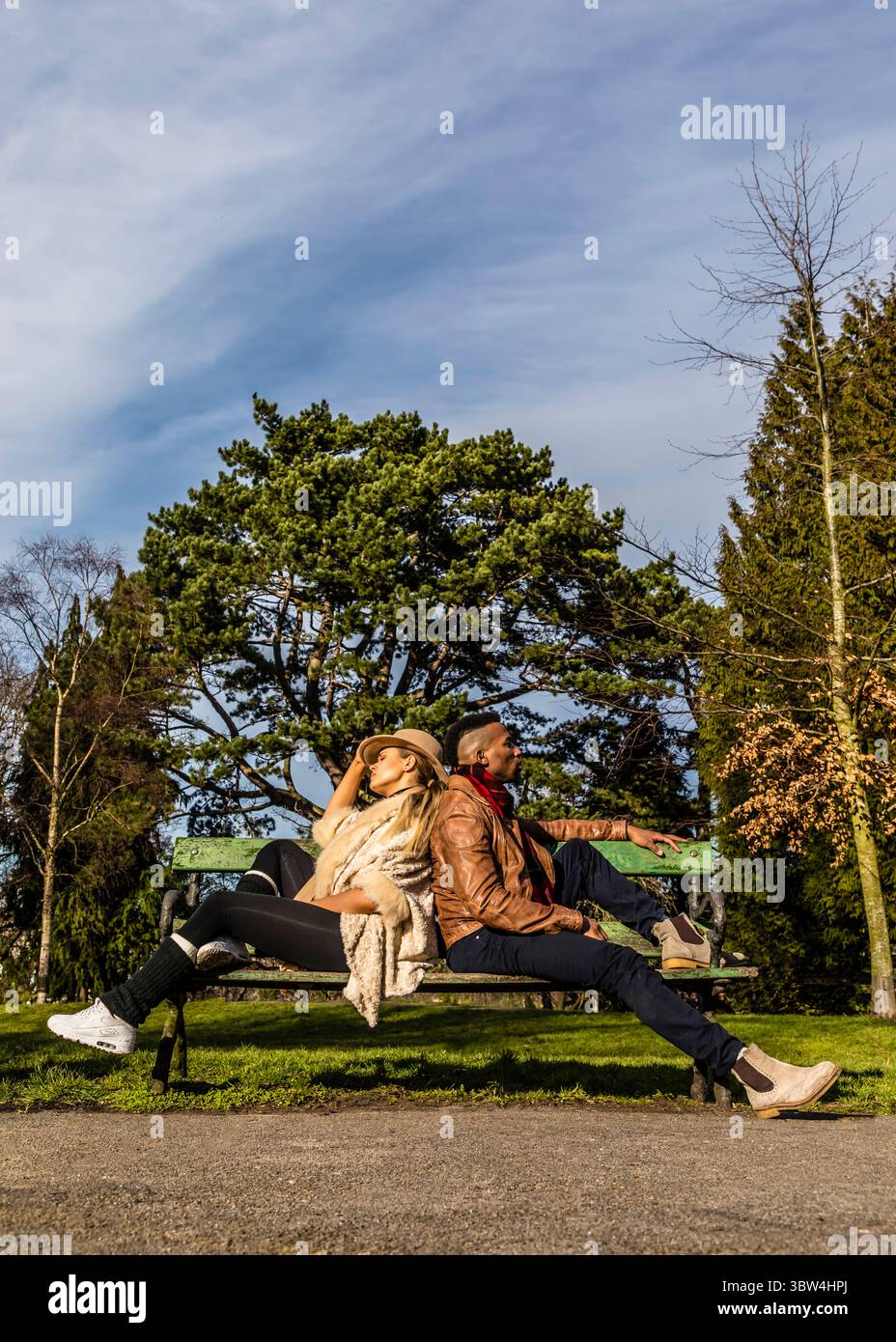 A couple is sitting back to back on a park bench. The woman is wearing ...