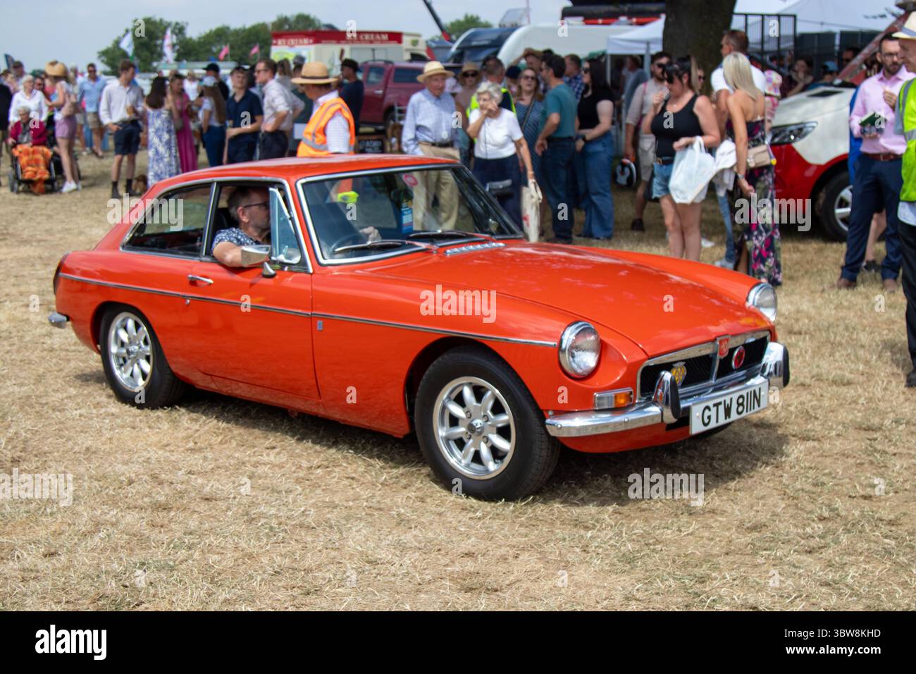 A Red MG B convertible at a UK country fair, driven past crowds during a classic car show ...