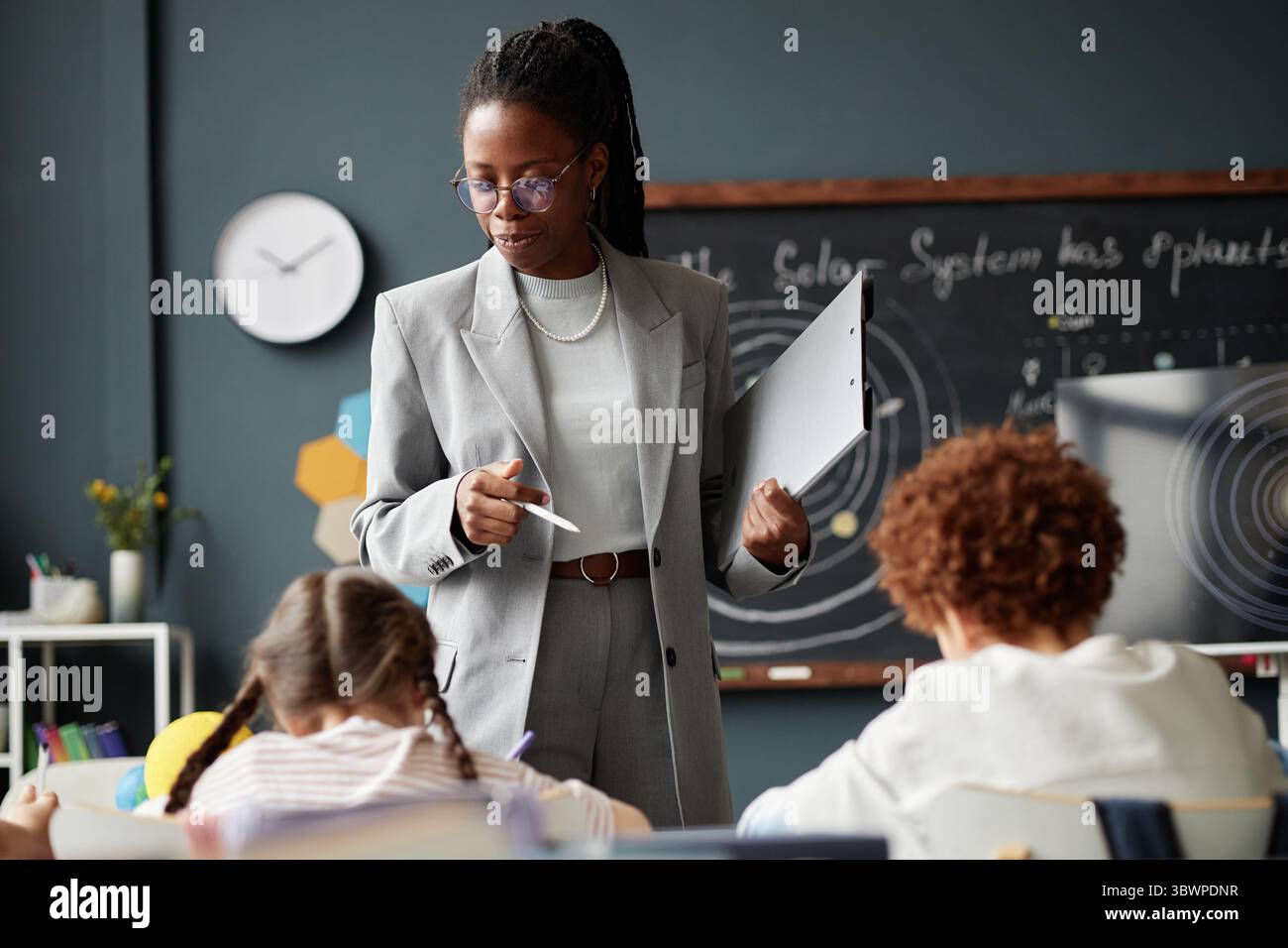 Black woman teacher standing in classroom holding clipboard and pen ...