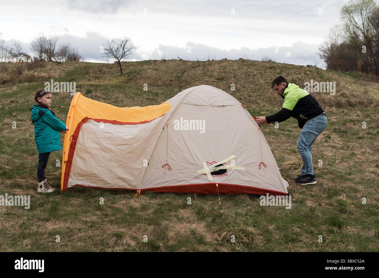 On a brisk day, a child in a teal jacket assists an adult in pitching a ...