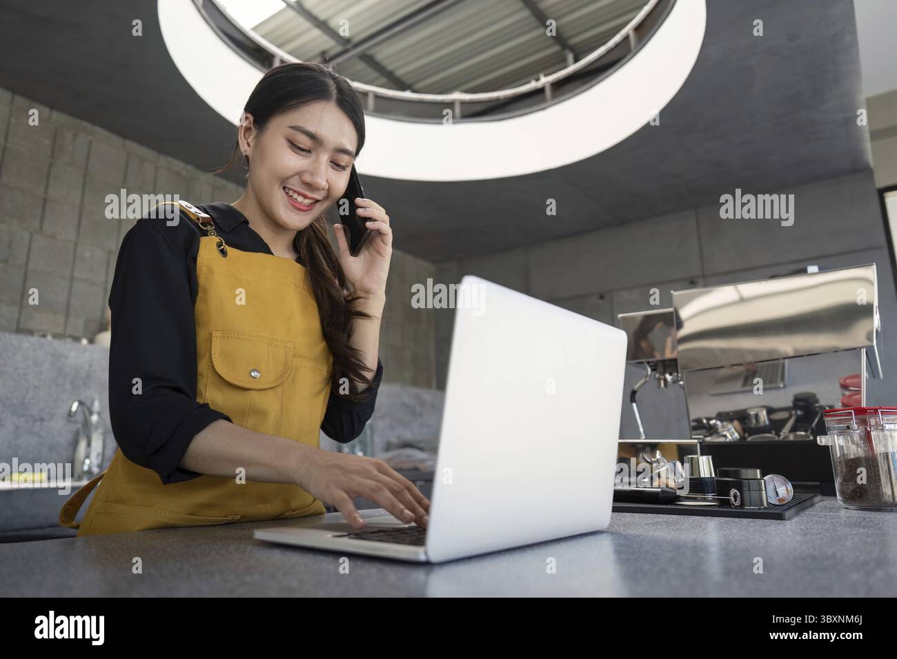 Smiling young woman barista in yellow apron multitasking with laptop ...
