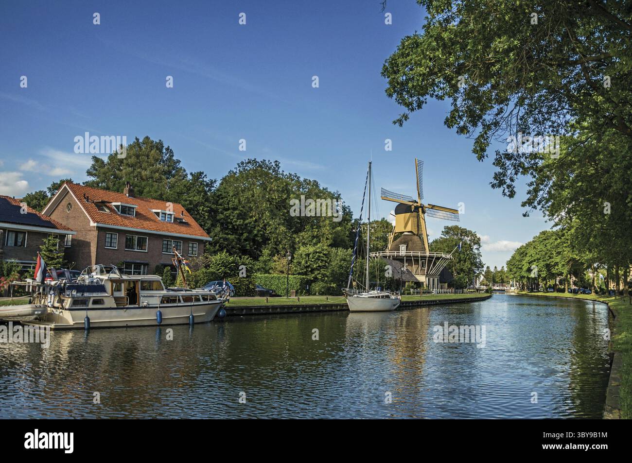 Boats and wooden yellow windmill next to wide tree-lined canal on the ...