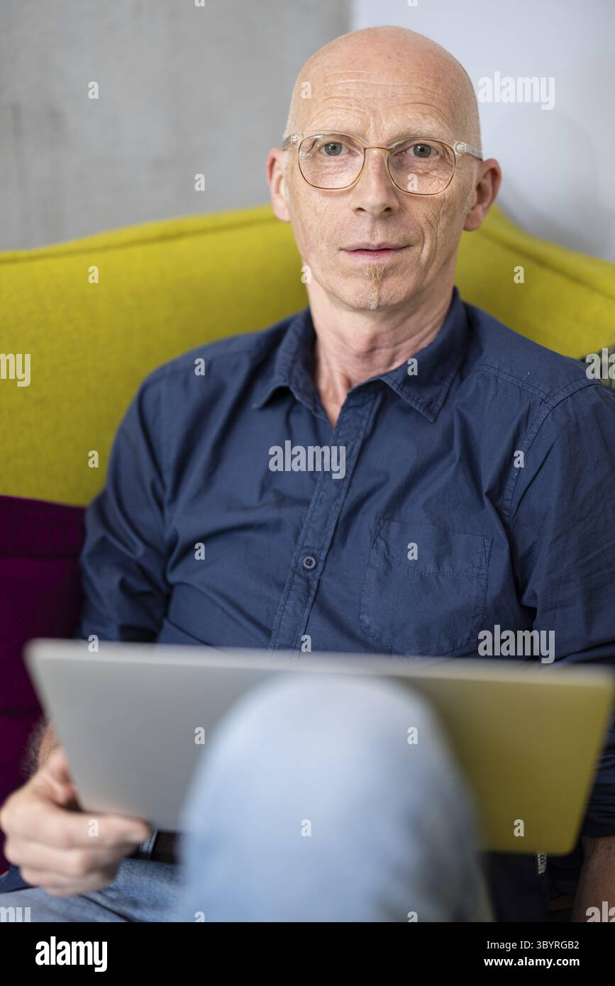 Elderly, bald man with glasses sits relaxed with his laptop on a yellow ...