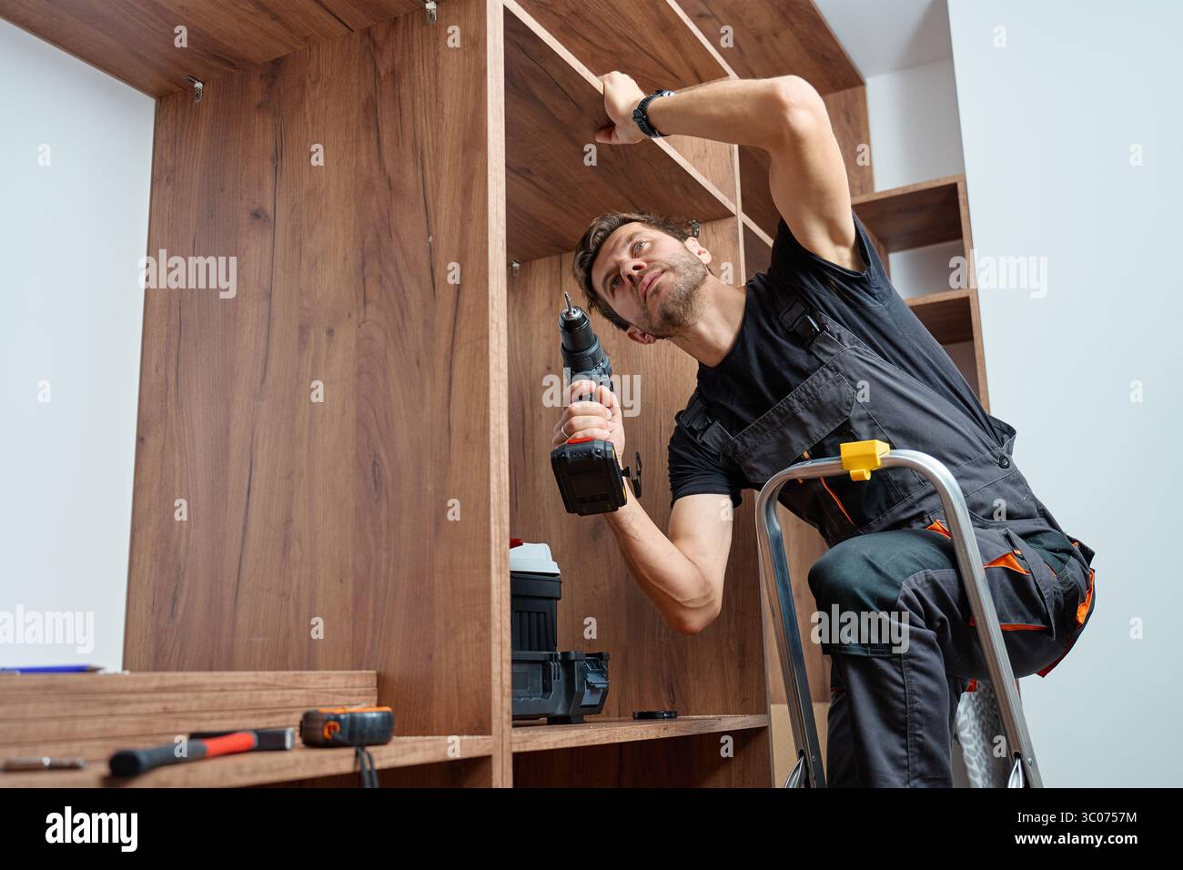 Man wearing work overalls assembling wooden wardrobe with drill and tools. Carpenter standing on ...