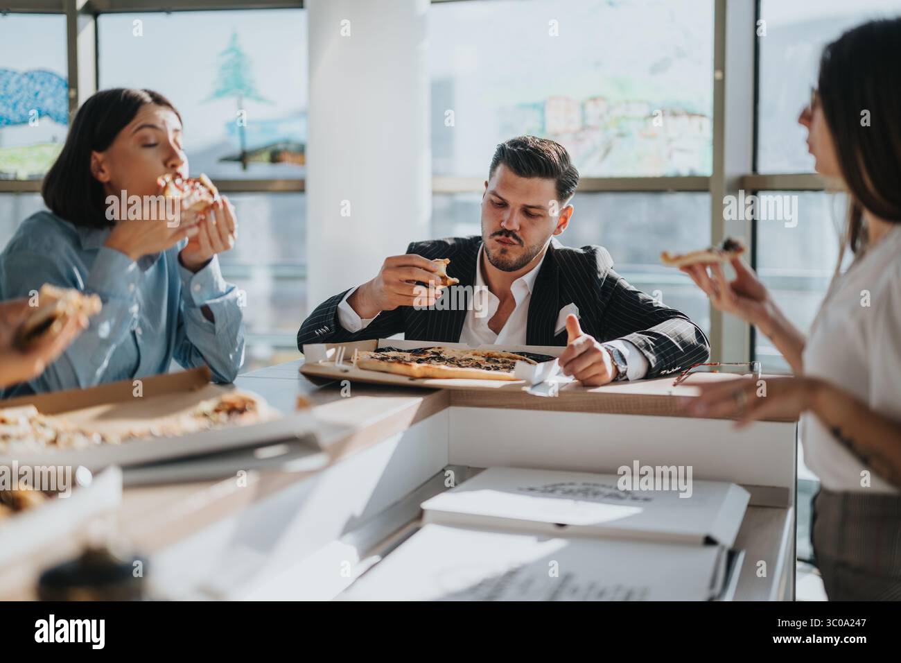 Office colleagues enjoying pizza during lunch break in sunlit workspace ...
