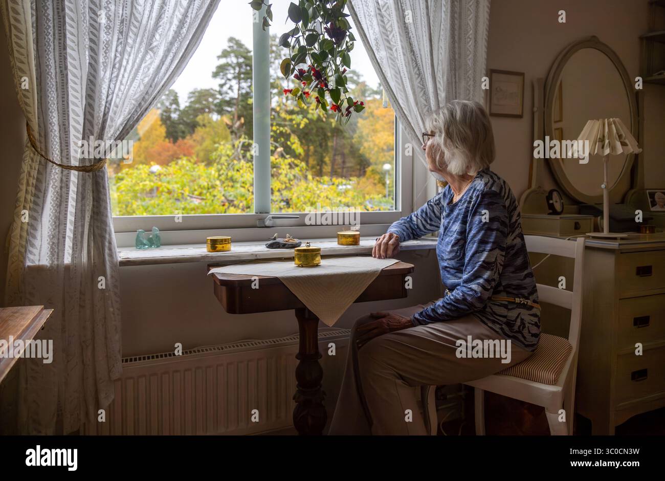 Elderly woman sitting alone by a window in a cozy home, looking out at ...