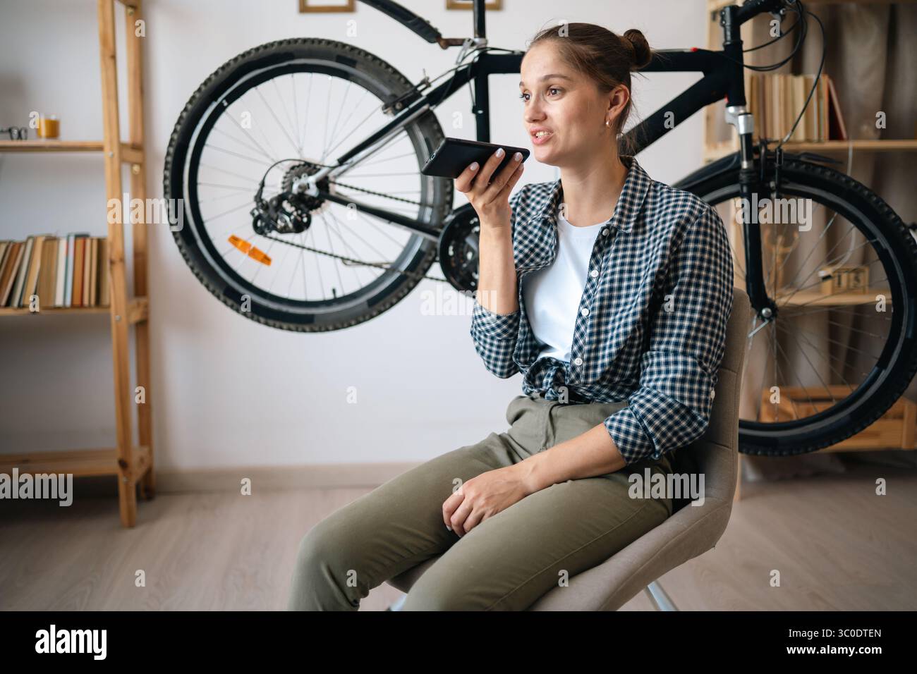 woman repairing bicycle at home using voice assistant and smartphone to send message and ask for ...