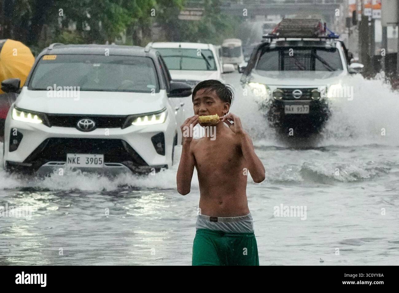 A boy eats corn in front of vehicles along a flooded road following ...