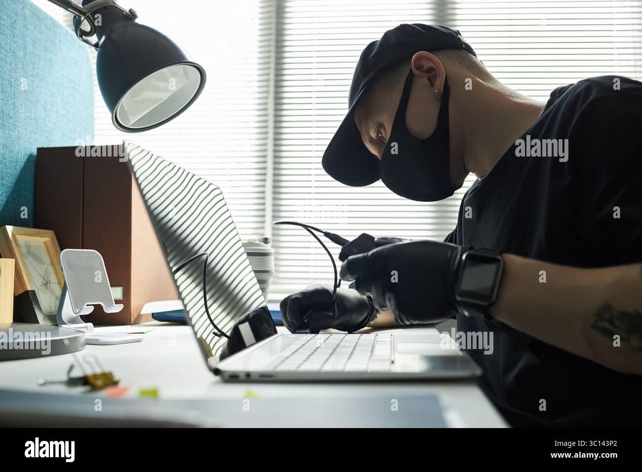 Male hacker wearing black cap and face mask sitting at desk in office ...