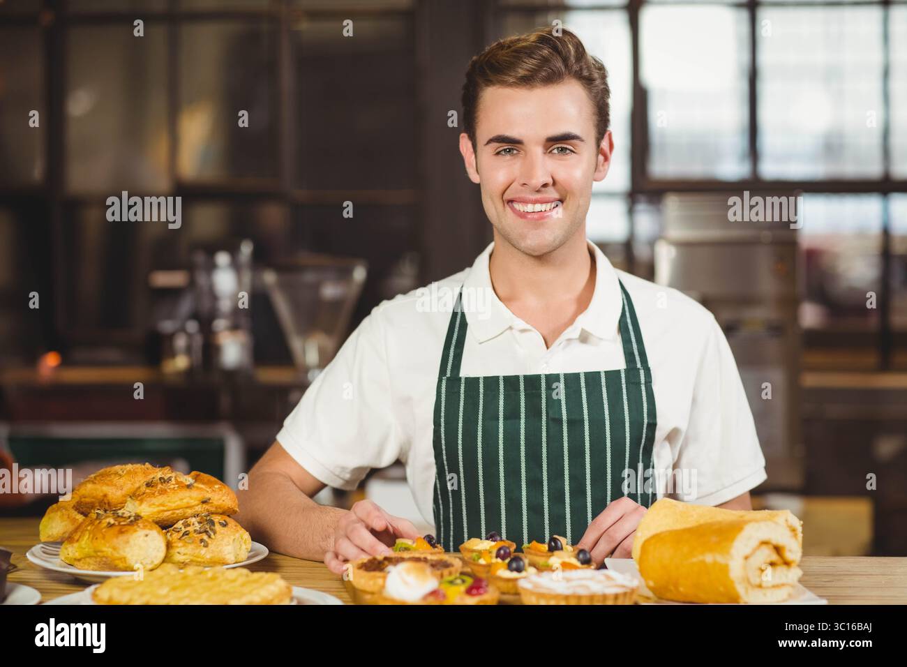 Male baker wearing green-striped apron arranging pastries on wooden ...