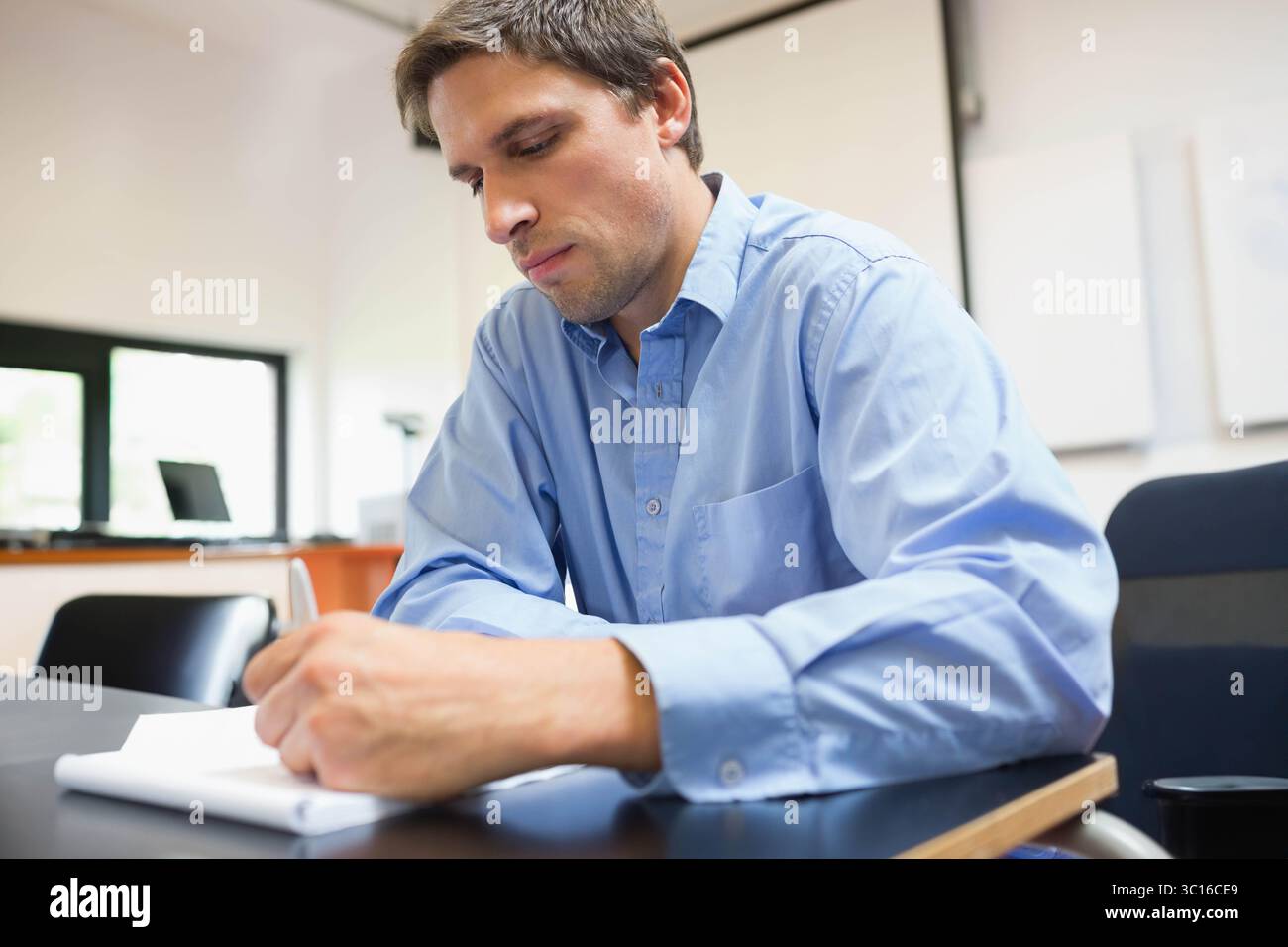 Male student writing notes in notebook at desk in classroom with laptop ...