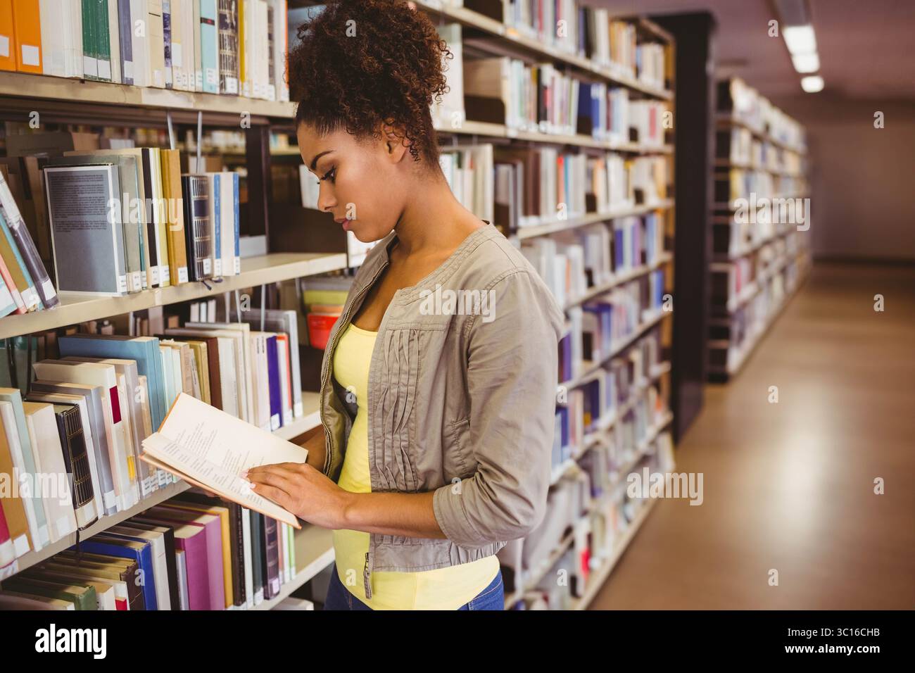 Woman reading open book in library aisle, with tall metal bookshelves ...