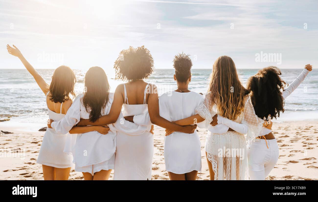 Back view of multiracial ladies in white dresses posing on beach and ...