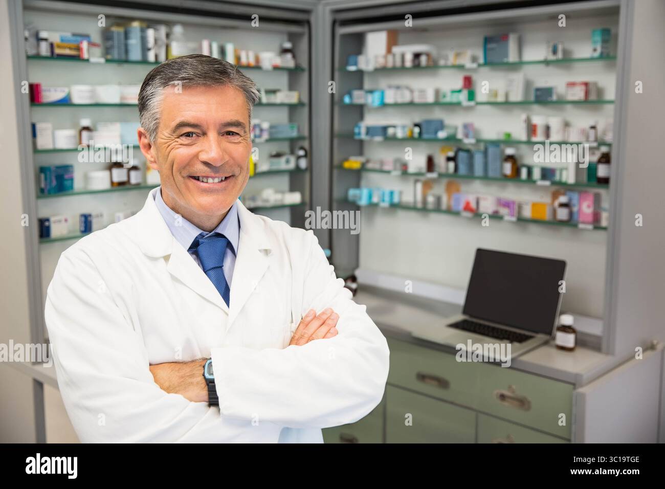 Senior male pharmacist wearing lab coat standing at counter with laptop ...