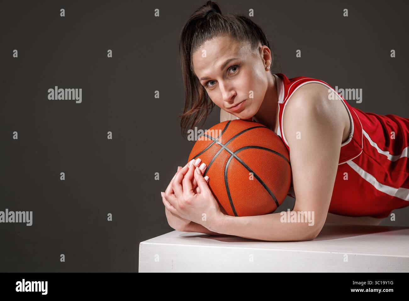 Confident female athlete in red basketball uniform holds ball in studio ...