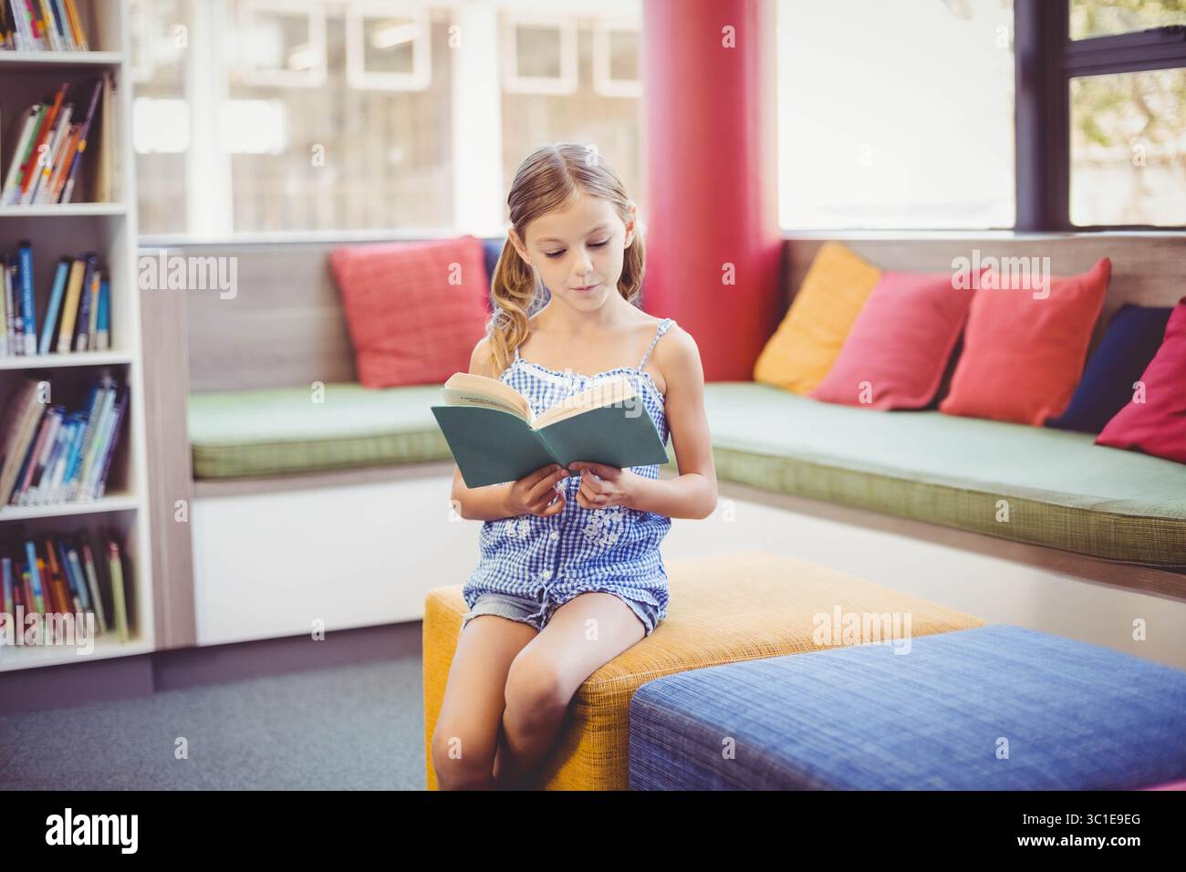 Female child reading open book while sitting on ottoman in library near ...