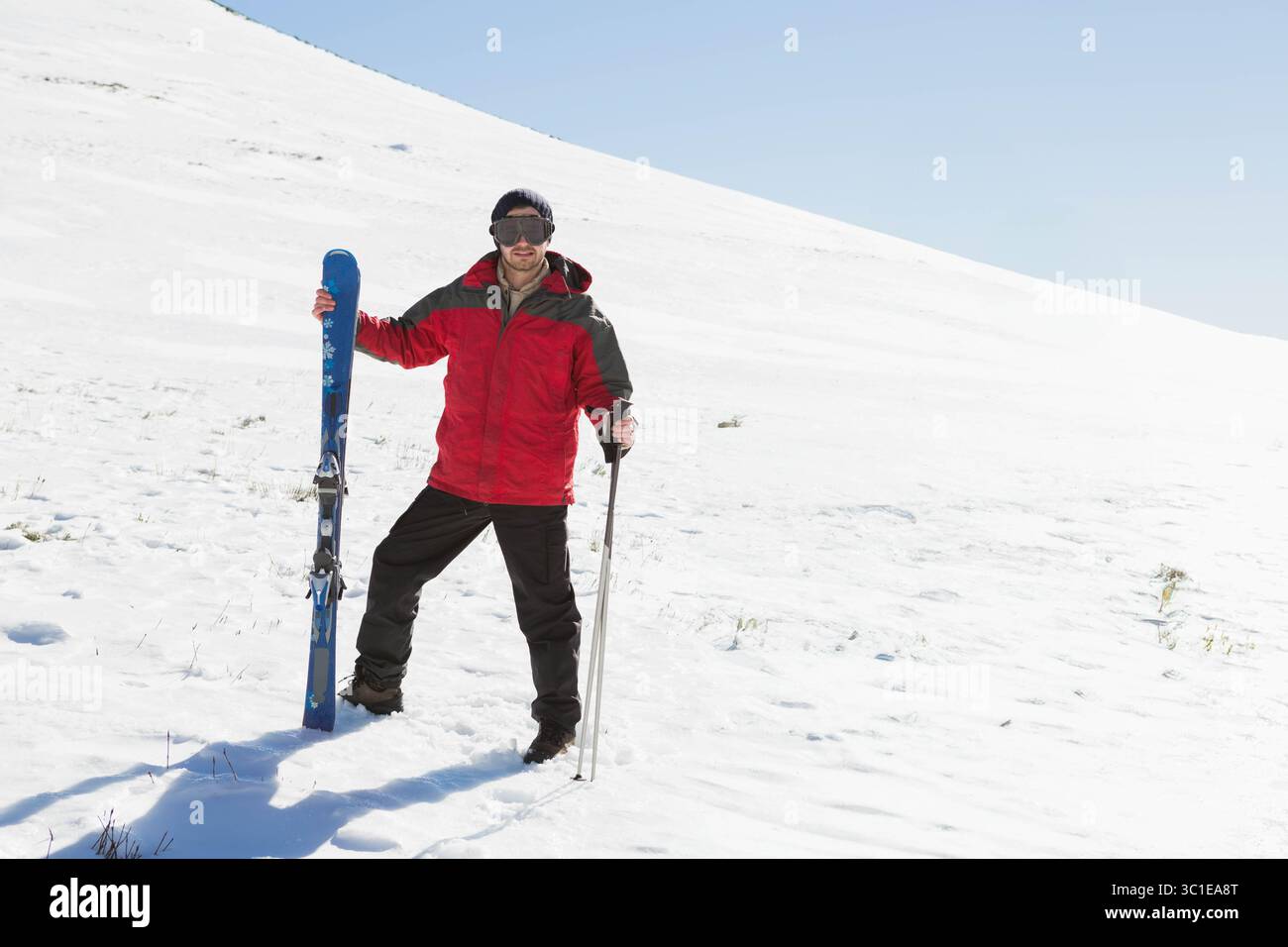 Skier standing on gentle snow slope wearing red jacket and holding blue ...