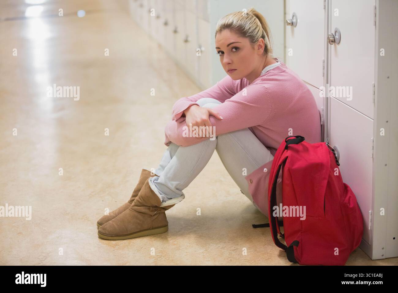 Teenage female student sitting in school hallway hugging knees by ...