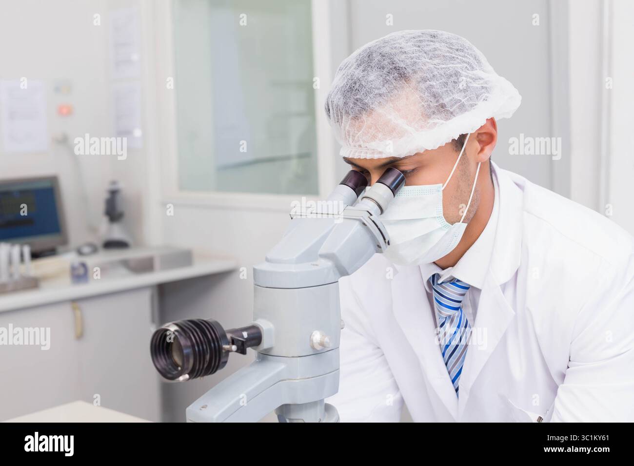 Lab technician peering through light microscope on bench, with computer ...
