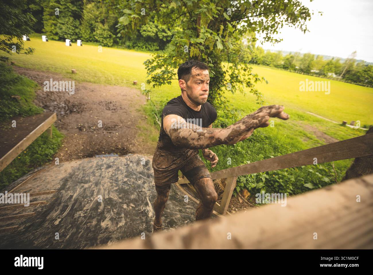Muddy man is scaling wooden obstacle wall on muddy ramp with field ...