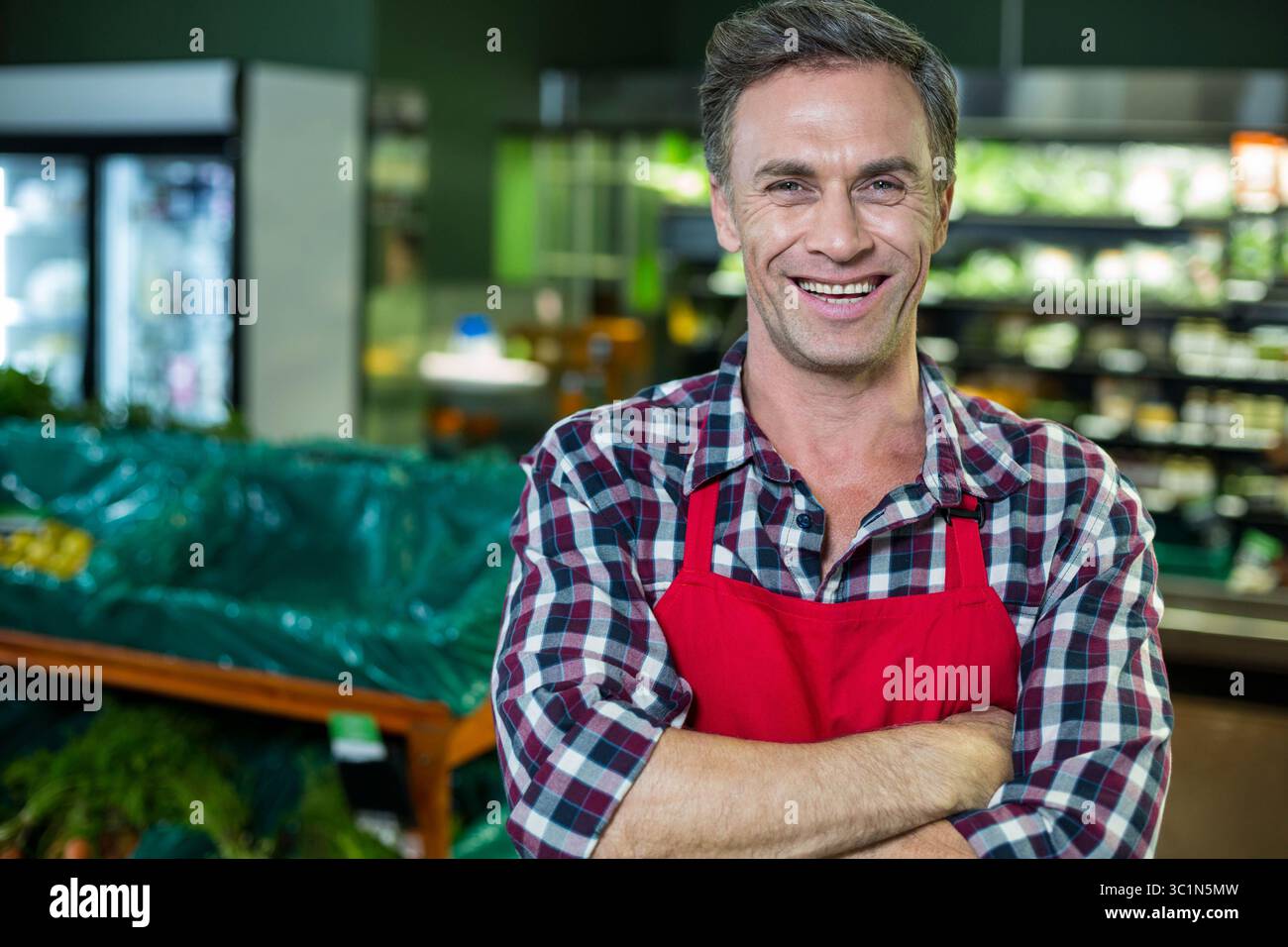 Man standing wearing red apron with arms crossed in produce section ...