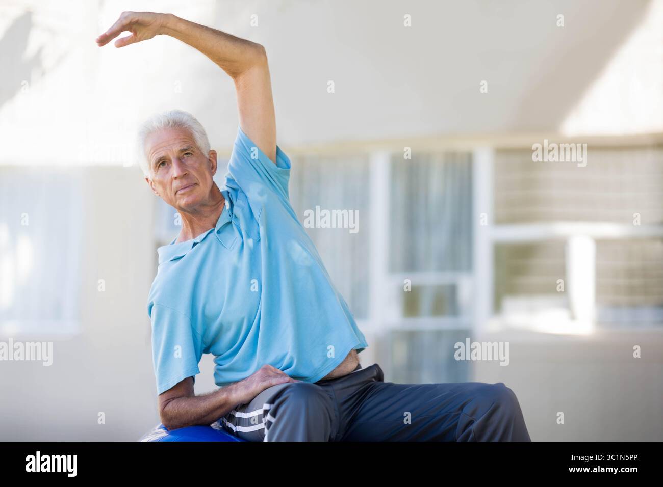 Senior man sitting on blue stability ball stretching left side on ...